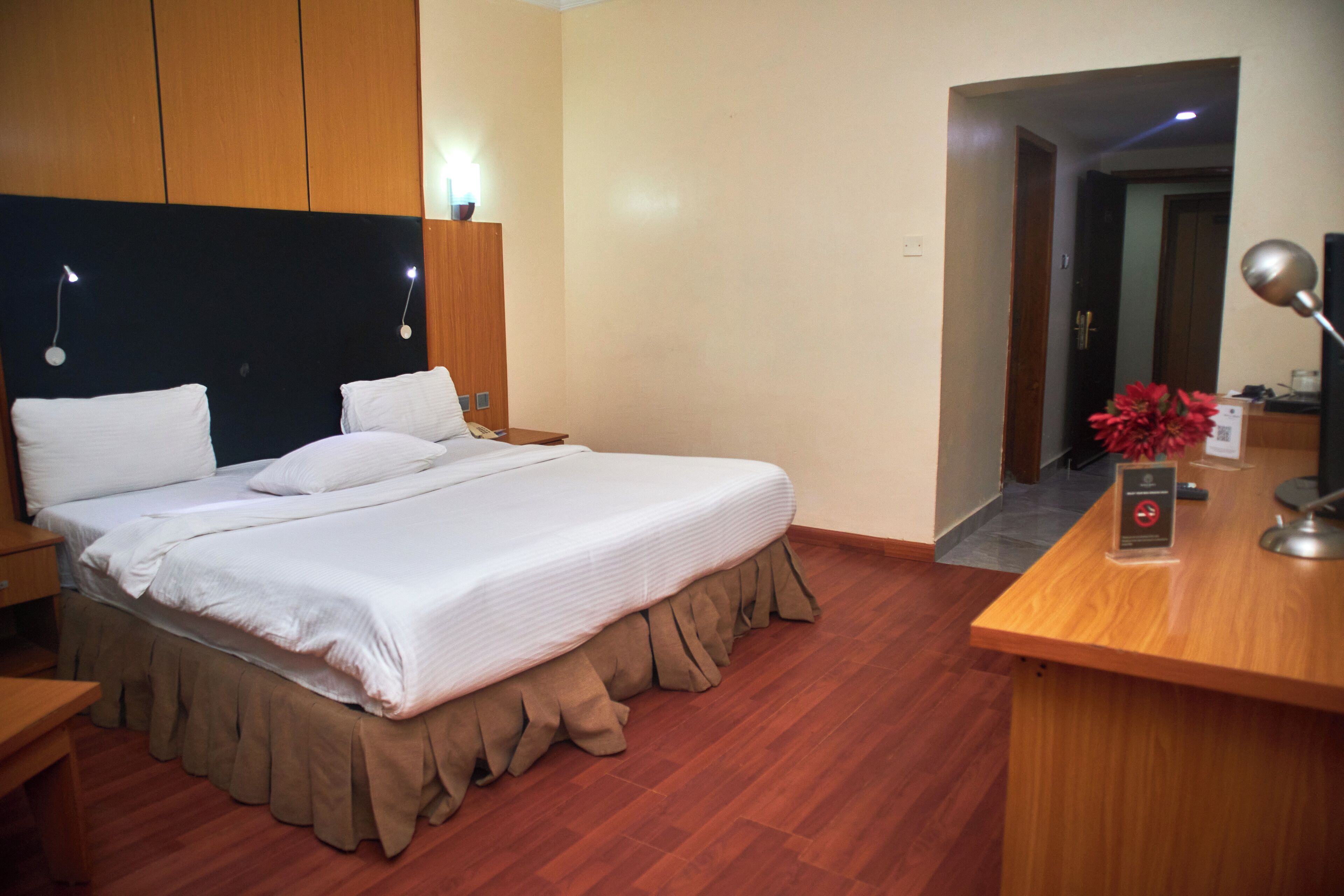 Quarto de hotel com uma cama grande, cabeceira de madeira, mesas de cabeceira, uma escrivaninha e uma pequena luminária de mesa.