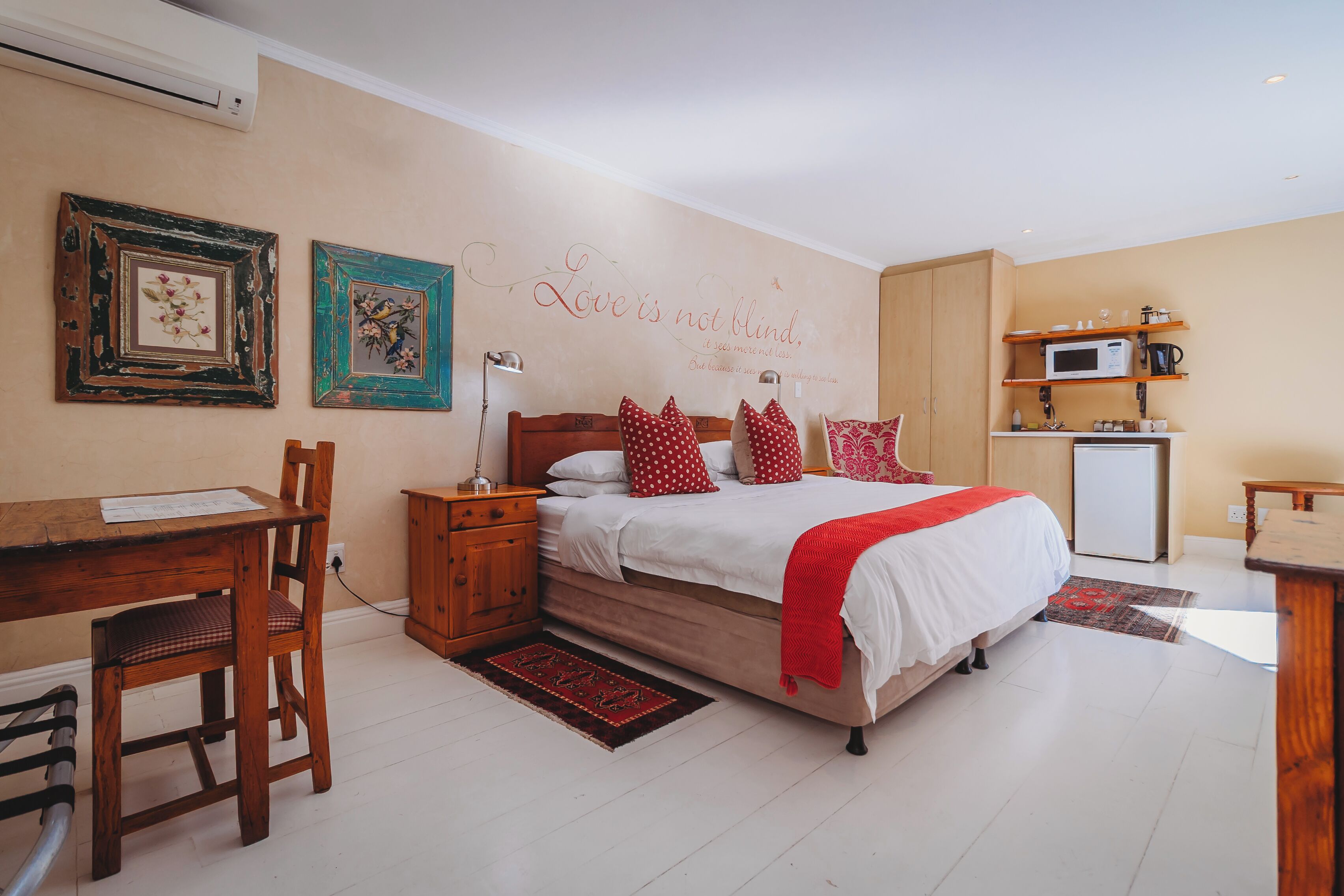 Quarto com cama, mesa de cabeceira, cadeira e pequena área de cozinha.