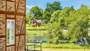 Um edifício de tijolos com janela e porta visíveis, um lago e uma casa vermelha ao fundo.