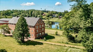 Um edifício vermelho grande e tradicional, com telhado inclinado, cercado por vegetação exuberante e um pequeno lago.