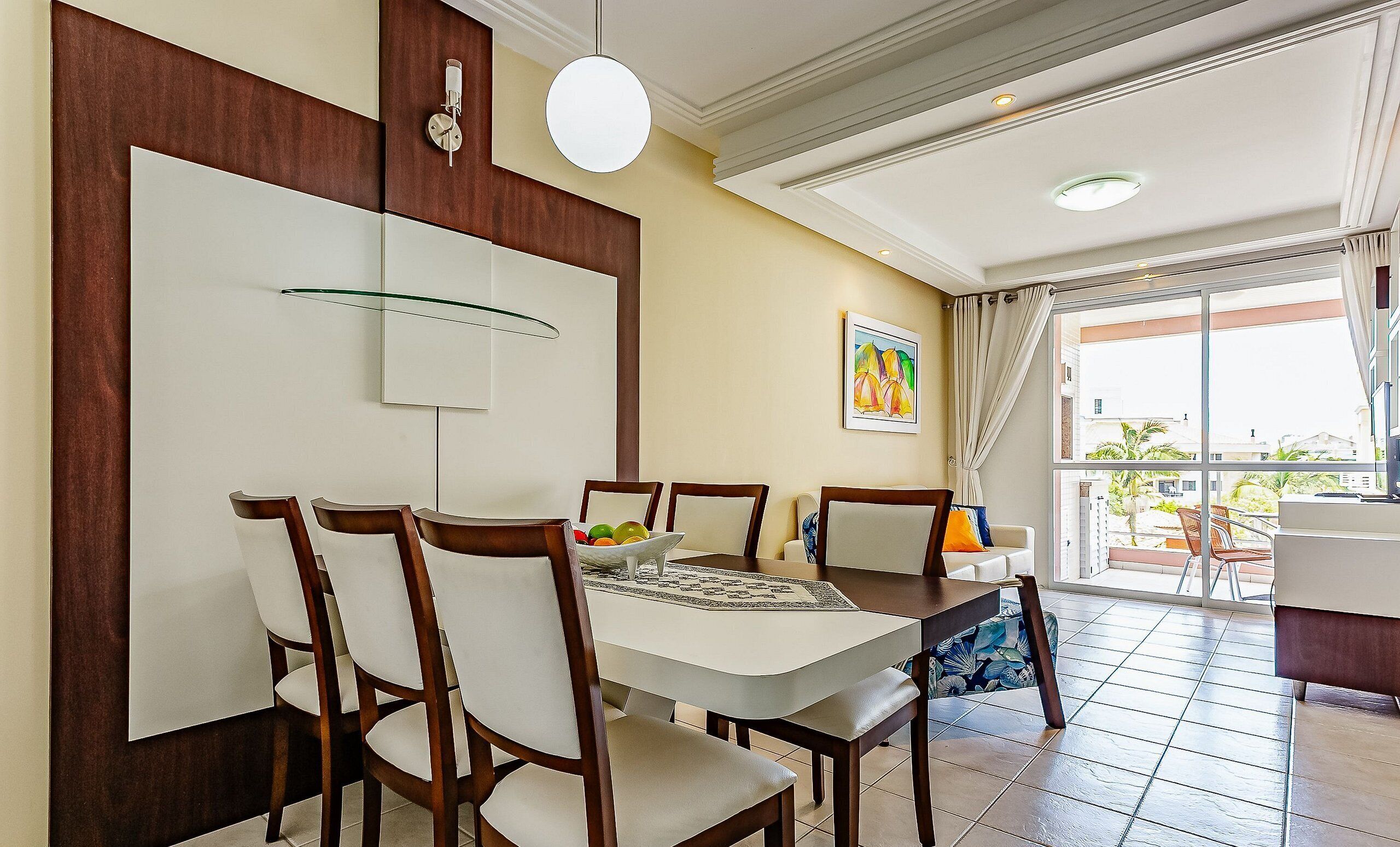 Uma sala de jantar com uma mesa posta para quatro pessoas, rodeada por cadeiras de madeira. Há um candeeiro fixado na parede acima da mesa e um quadro na parede.