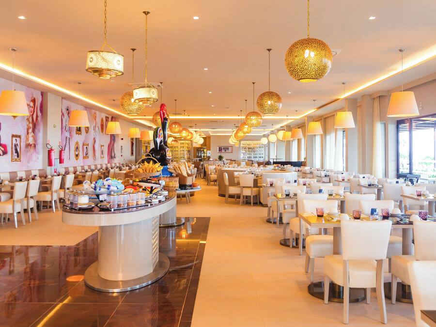Ampla área de refeições de hotel, com várias mesas e cadeiras, um buffet central e grandes janelas que permitem entrada de luz natural.