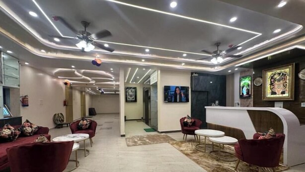 Um hall de hotel moderno com balcão de recepção, área de estar com cadeiras vermelhas e mesas brancas, e teto com iluminação embutida e um ventilador.