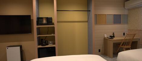 Quarto de hotel com uma cama, escrivaninha, cadeira, televisão, mini-geladeira e prateleira com chaleira.