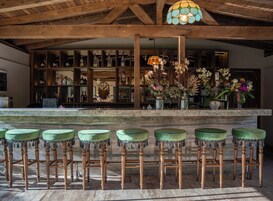 Uma área de bar com banquetas de madeira, balcão de mármore e diversos frascos e objetos decorativos.