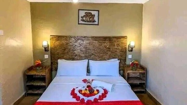 Quarto de hotel com uma cama grande, cabeceira de madeira, duas mesas de cabeceira com abajures e um quadro pendurado na parede.