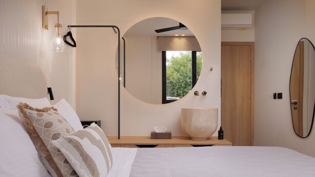 Quarto moderno com uma cama grande, um espelho redondo, uma mesa de cabeceira com abajur e vista para o verde pela janela.