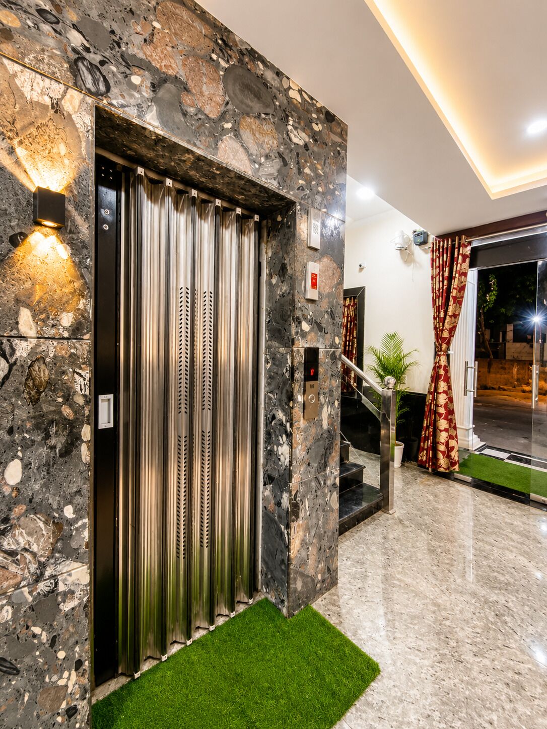 Um elevador com fachada de pedra, uma escadaria com cortinas vermelhas e uma área com grama sintética verde.