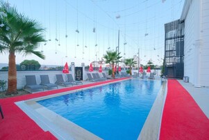 Uma piscina com bordas de carpete vermelho, cadeiras de descanso, palmeiras e um edifício com fachada moderna.