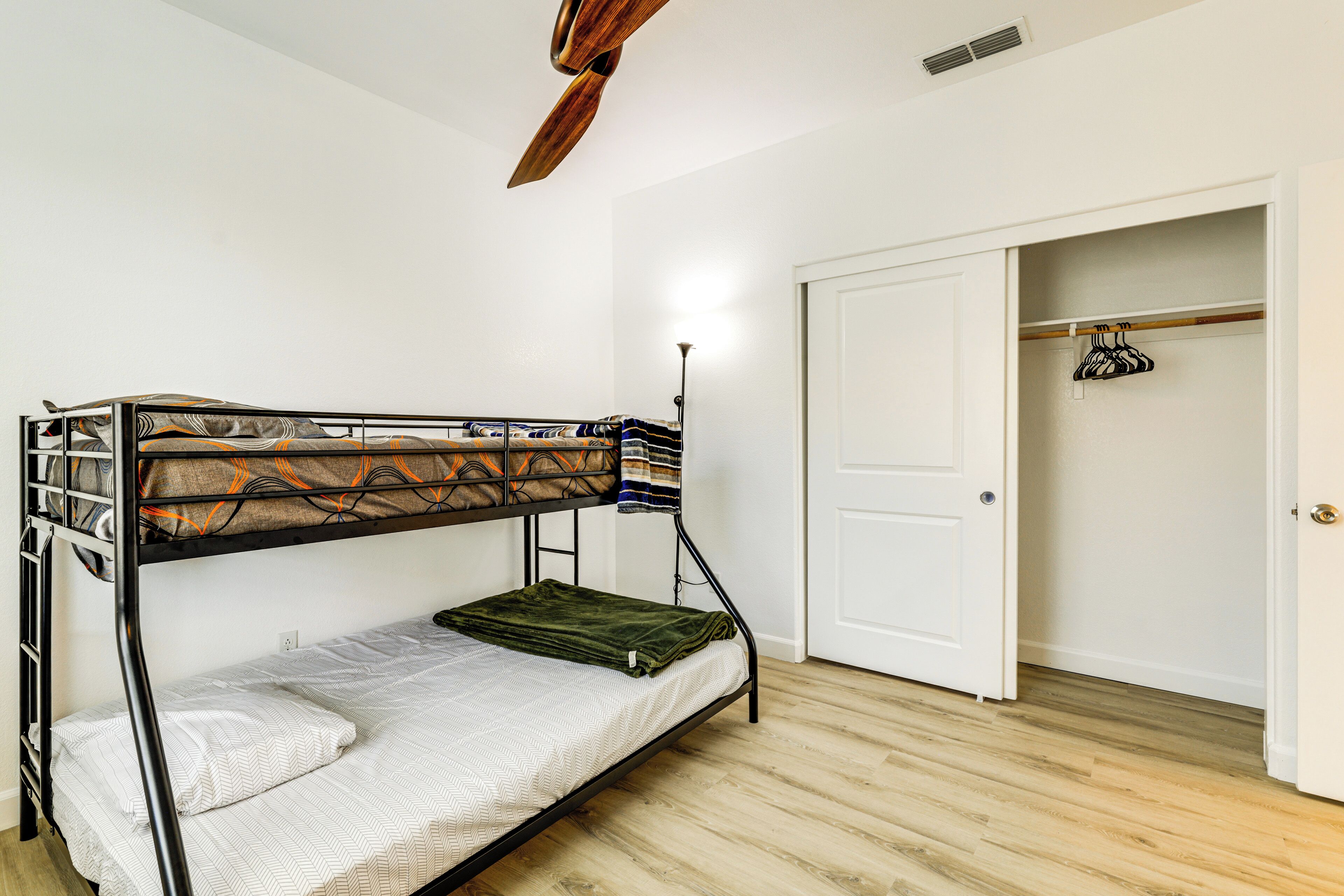 Uma cama beliche com um cobertor estampado, um ventilador de teto de madeira e uma porta branca com um porta-roupas.