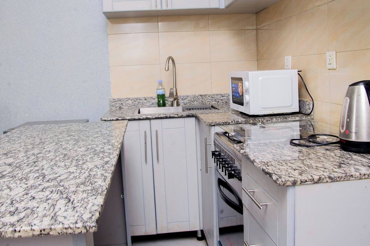 Uma cozinha compacta com armários brancos, bancada de granito, micro-ondas, chaleira e pia.