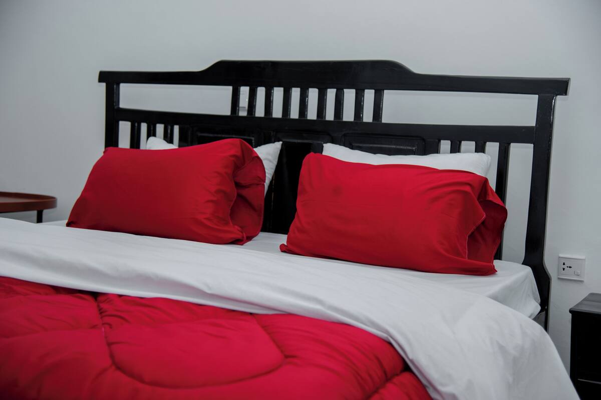 Uma cama com cabeceira preta, roupa de cama branca e duas almofadas vermelhas.