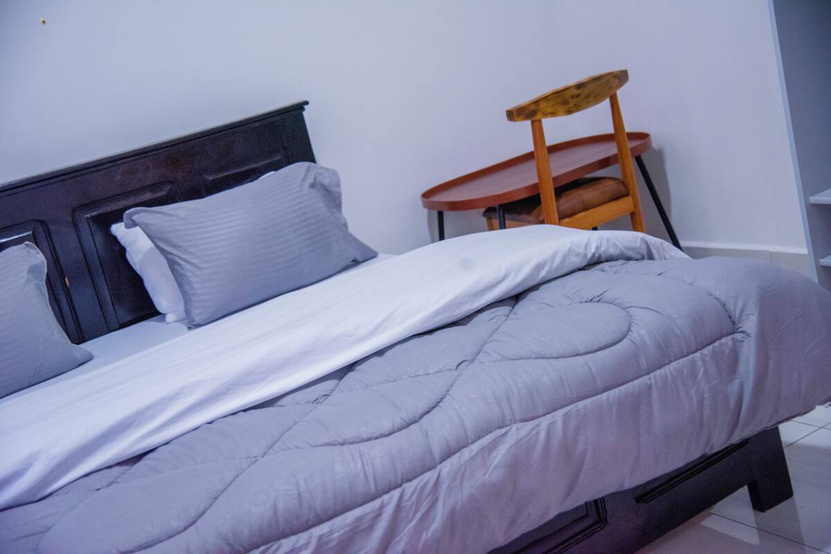 Uma cama bem arrumada com um edredom cinza e lençóis brancos, uma cadeira de madeira e uma cabeceira escura.