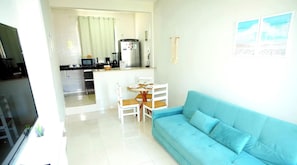 Sala de estar com sofá azul, área de jantar com mesa e cadeiras, cozinha com geladeira e micro-ondas, e vista para o exterior.
