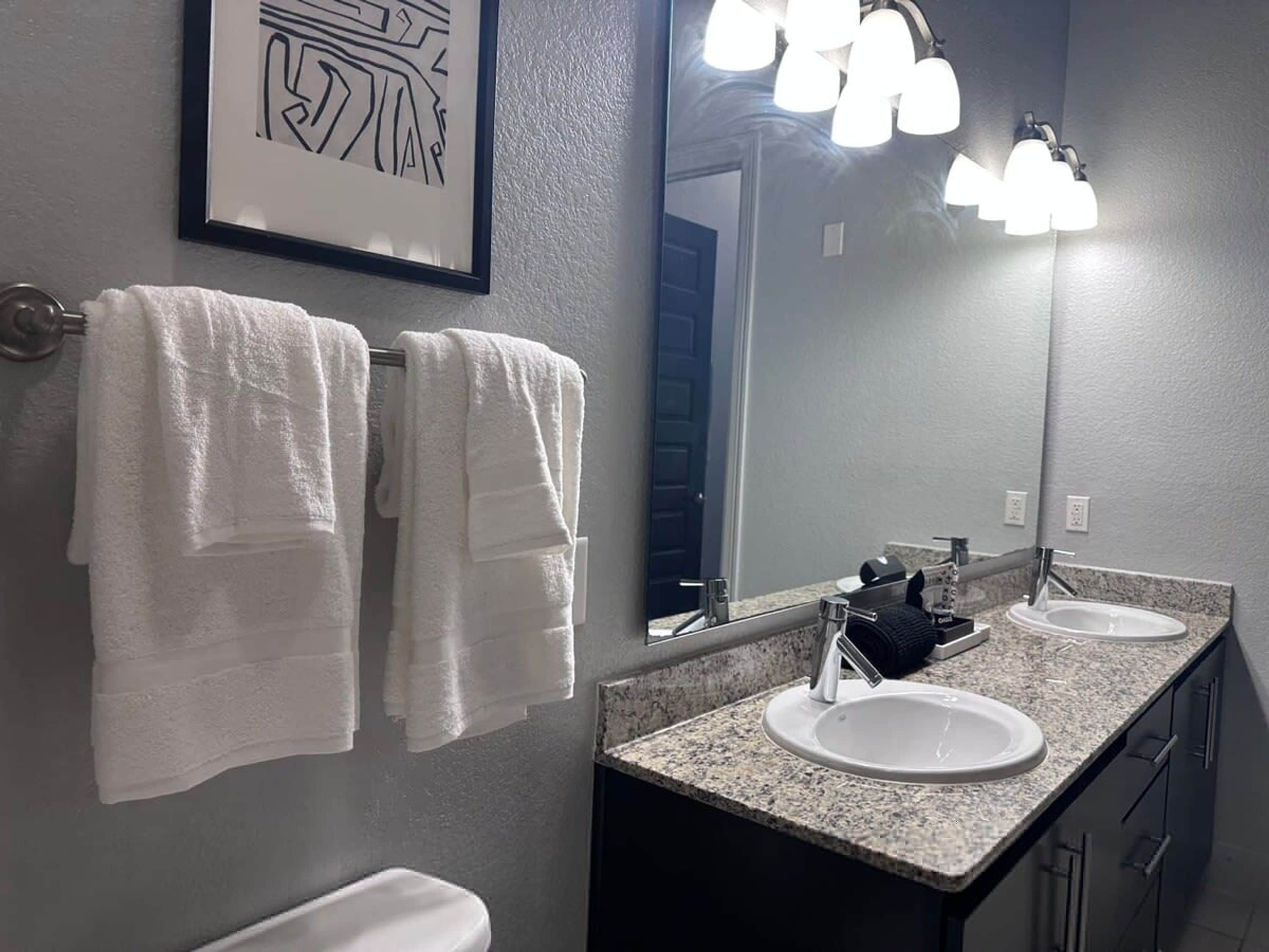 Uma casa de banho com duas toalhas brancas penduradas numa estante, um espelho grande com uma luminária e uma bancada de granito com uma pia.