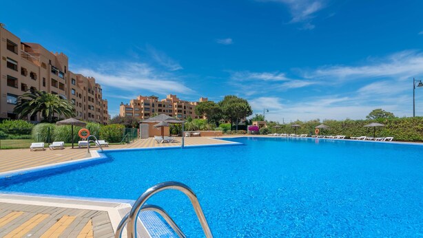 Uma piscina com água azul cristalina, rodeada por espreguiçadeiras e guarda-sóis, e um edifício com varandas ao fundo.