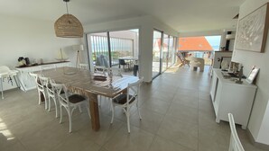 Ampla sala de estar com uma grande mesa de jantar de madeira, cadeiras brancas e luminária pendente.
