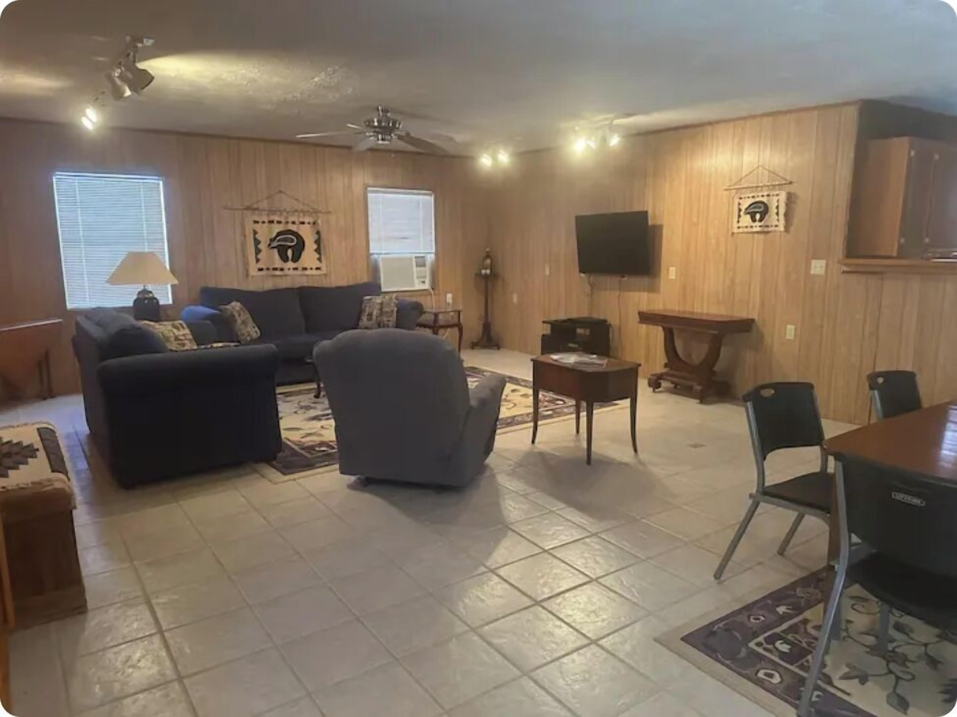 Uma sala de estar com sofá, poltronas, mesa de centro e televisão. As paredes são de madeira e há um ventilador de teto.