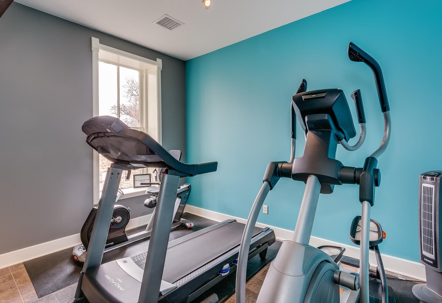 Uma academia em casa com esteira, elíptica e bicicleta ergométrica, uma janela com vista e uma parede azul.