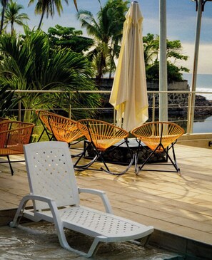 Área de piscina com espreguiçadeiras e mesa, com vista para um corpo d'água e palmeiras ao fundo.