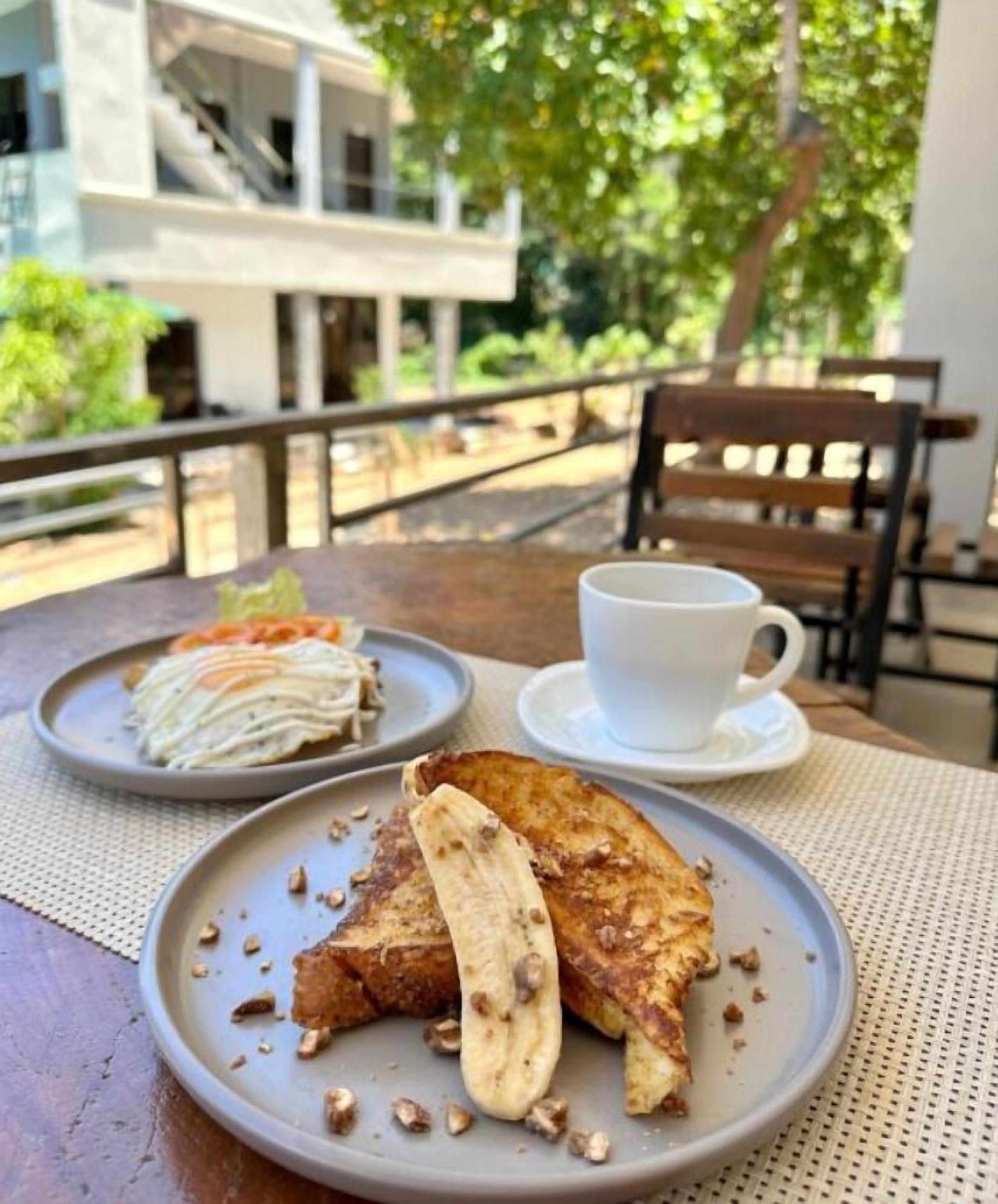 Mesa ao ar livre com torradas e banana, panquecas e uma xícara branca.