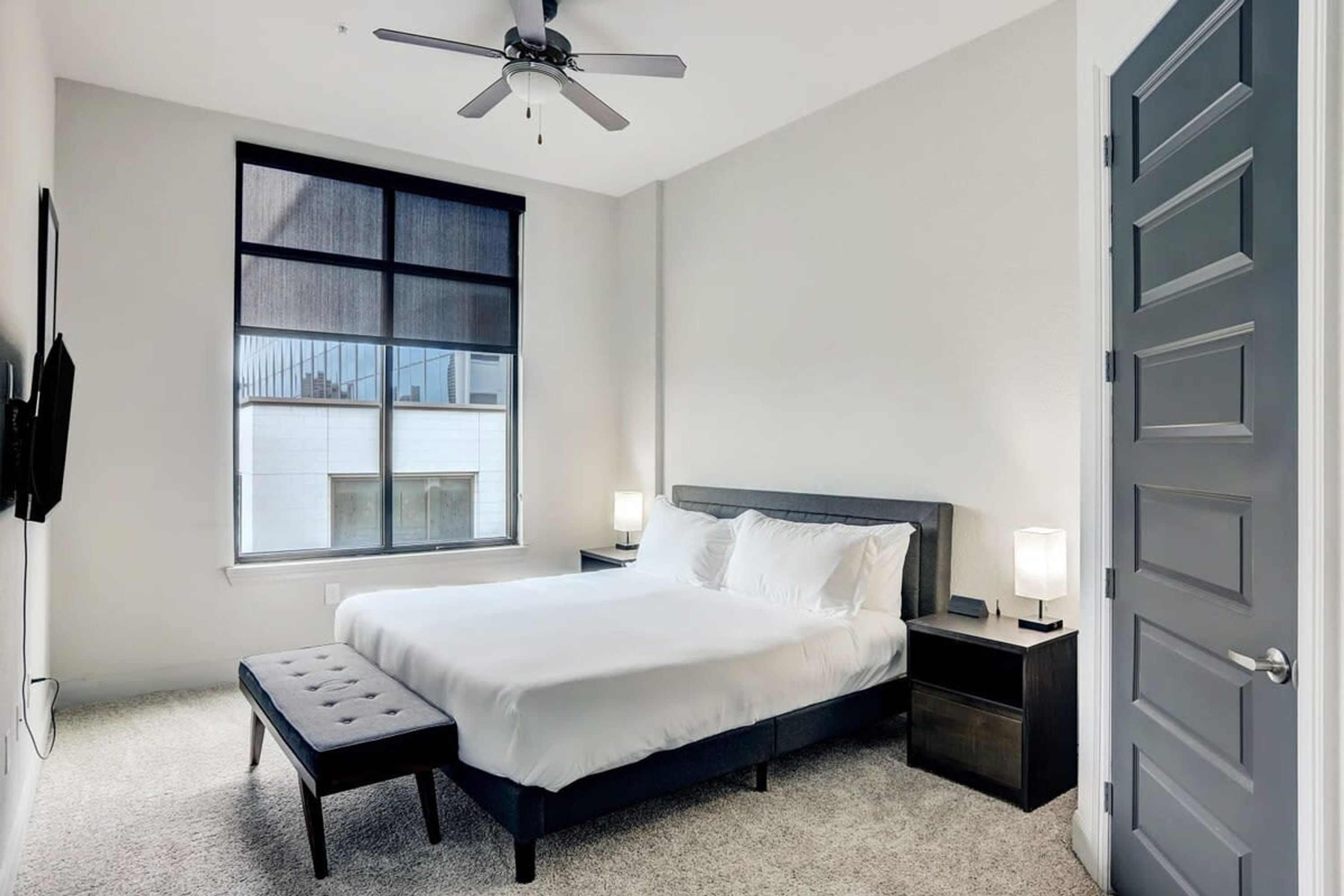 Quarto moderno com uma grande janela, uma cama, mesa de cabeceira com abajur, um banco e um ventilador de teto.
