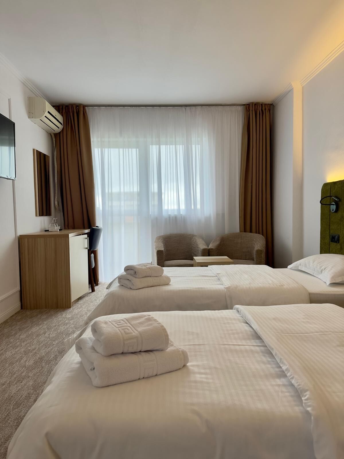 Quarto de hotel com duas camas, uma escrivaninha, uma cadeira e uma janela grande com cortinas.