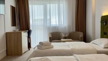 Quarto de hotel com duas camas, uma escrivaninha, uma cadeira e uma janela grande com cortinas.