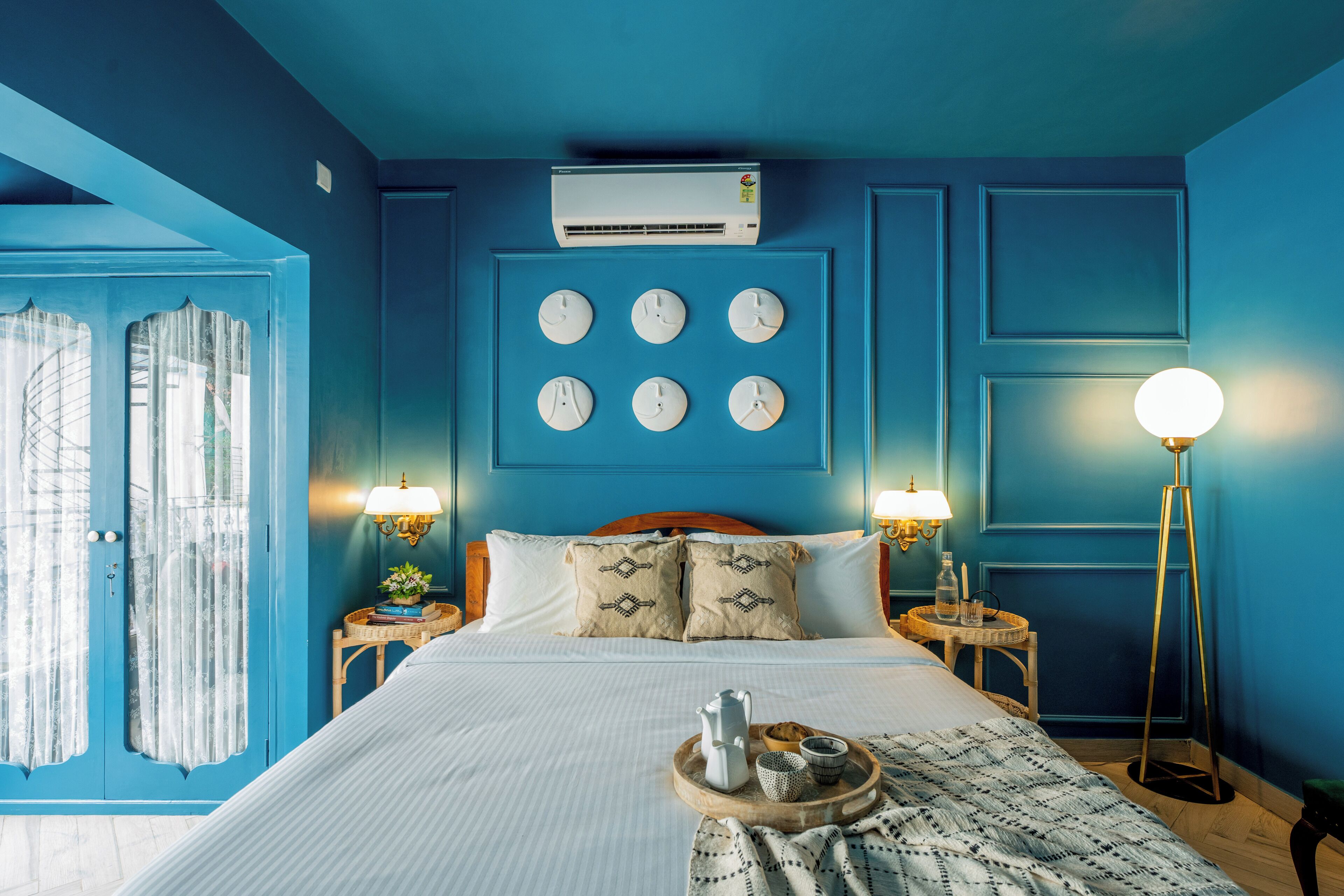 Quarto com cama, duas mesas de cabeceira, ar condicionado, relógio na parede, luminária de chão e janela com cortinas.