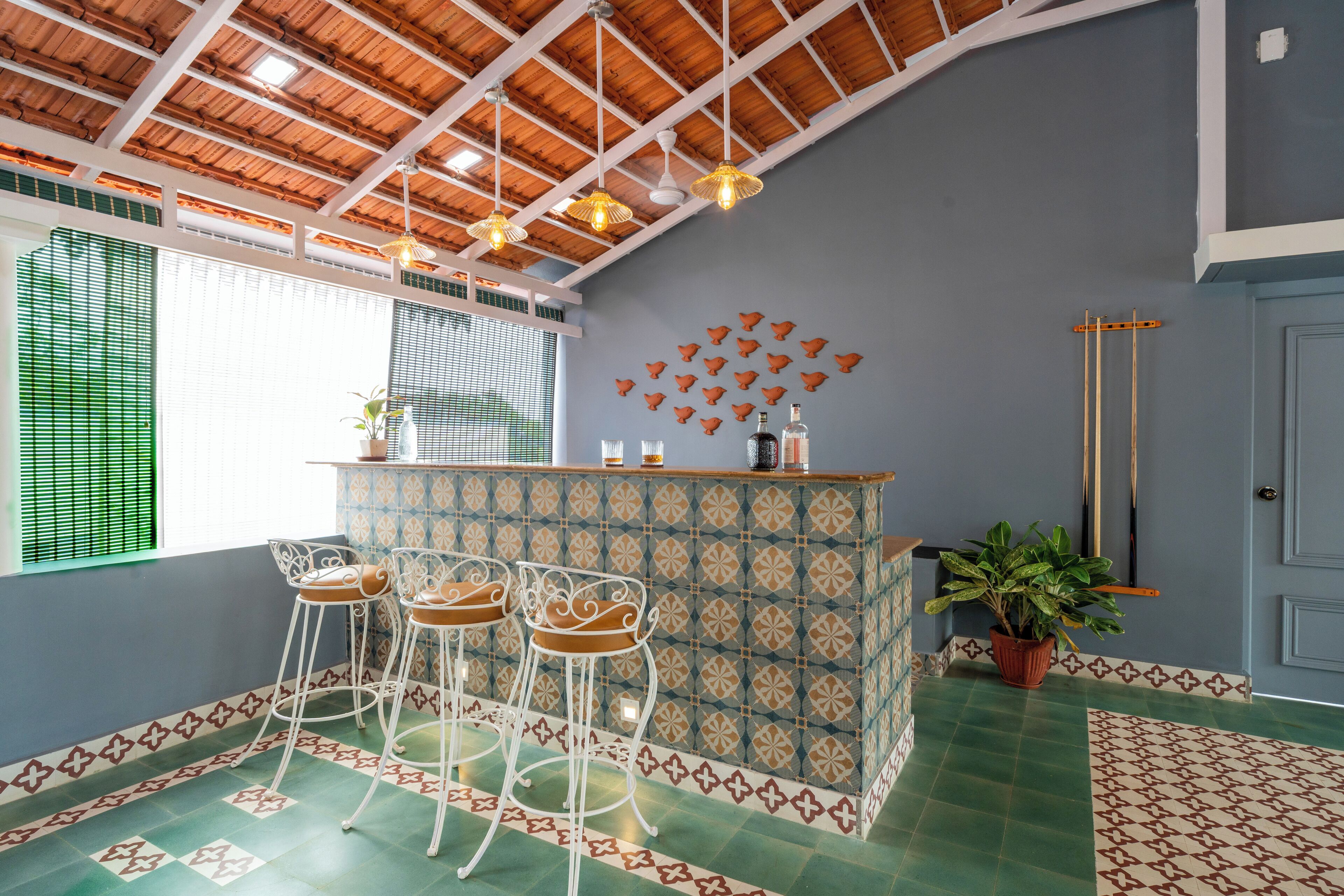 Uma área de bar com parede de azulejos estampados, três banquetas brancas e teto com vigas de madeira e luminárias pendentes.