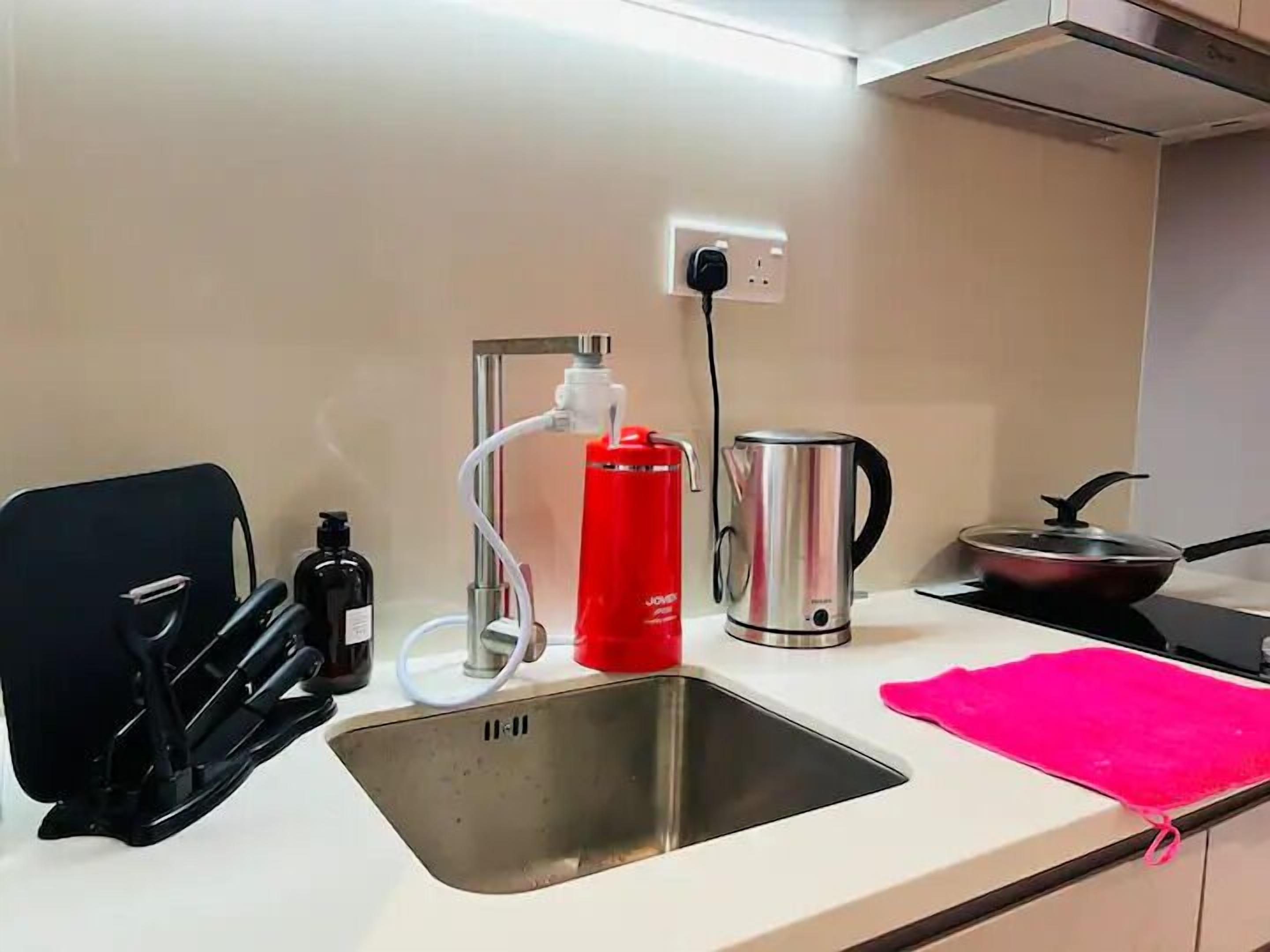 Cozinha com pia de aço inoxidável, dispenser de água vermelho, chaleira e uma frigideira no fogão.
