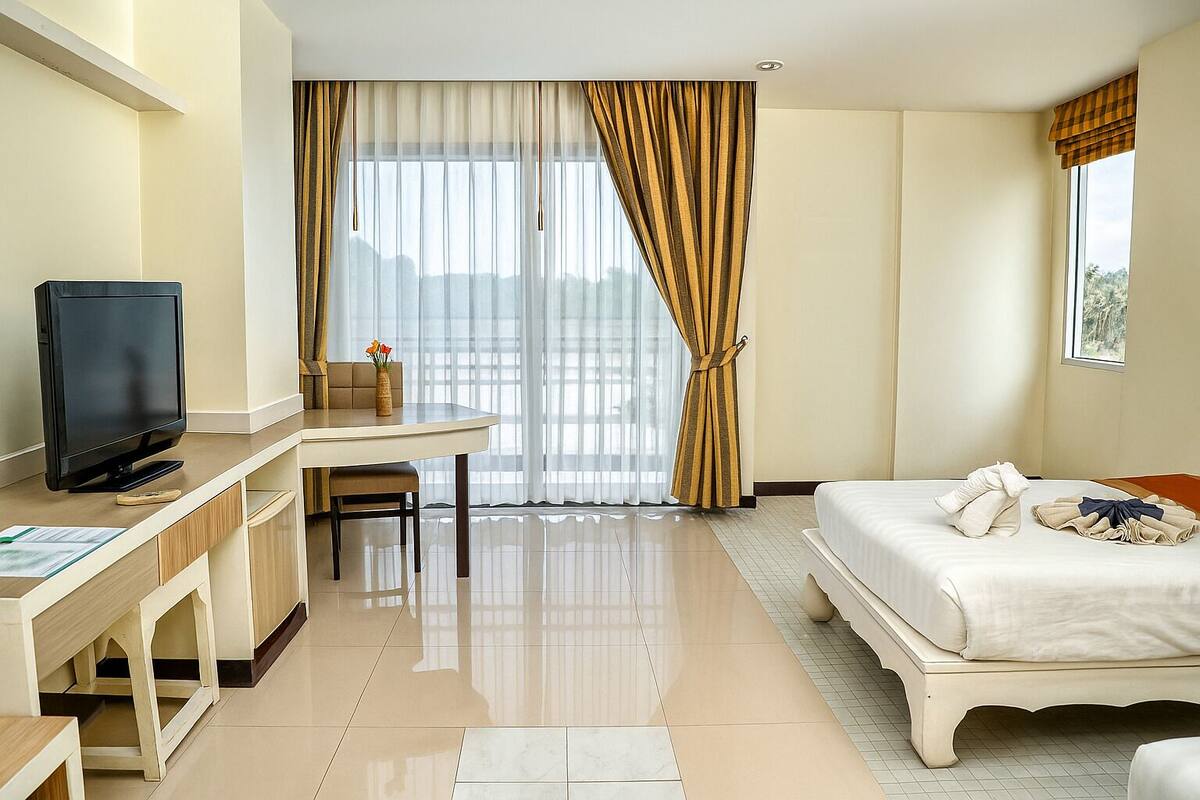 Quarto de hotel com cama, escrivaninha com televisão, cadeira e vista para o exterior através de amplas janelas.