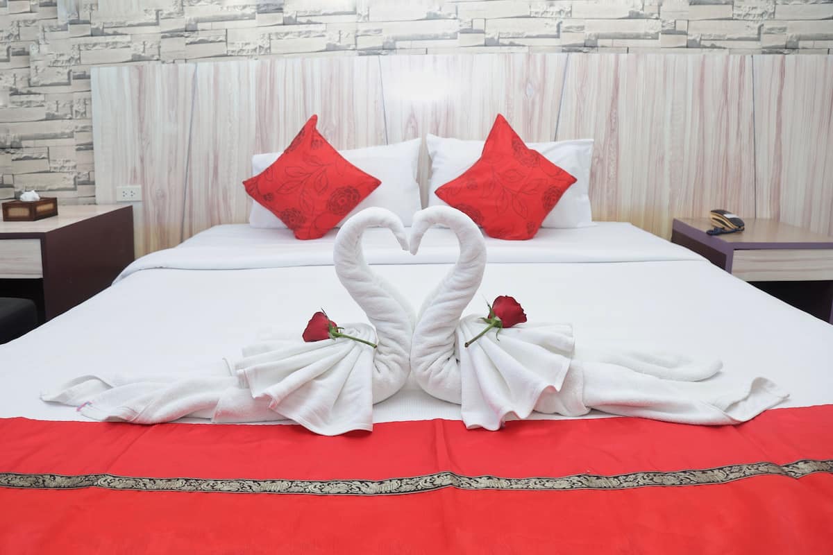 Quarto de hotel com uma cama bem arrumada, duas almofadas vermelhas e uma toalha disposta em formato de coração com rosas.