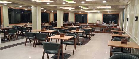 Um restaurante amplo, com várias mesas e cadeiras, tetos altos com iluminação embutida e piso revestido de azulejos.