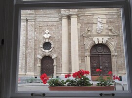 Fachada de edifício com duas portas grandes, uma estátua acima da porta esquerda e um canteiro de flores em primeiro plano.