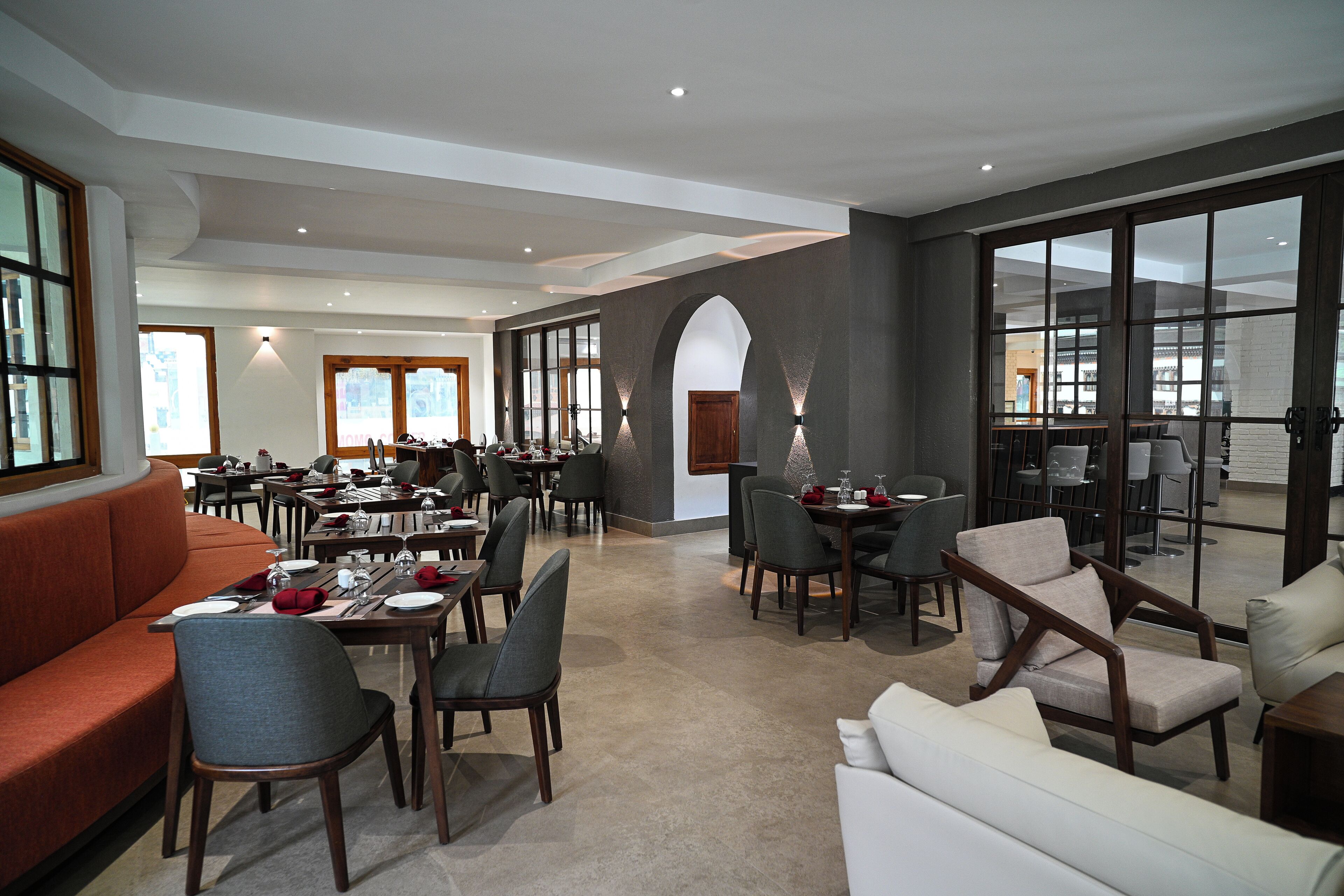 Uma sala de jantar ampla, com várias mesas e cadeiras, um sofá vermelho e grandes janelas que proporcionam luz natural.