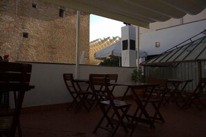 Um pátio externo com mesas e cadeiras de madeira, um muro de tijolos e vista para um edifício moderno com telhado inclinado.