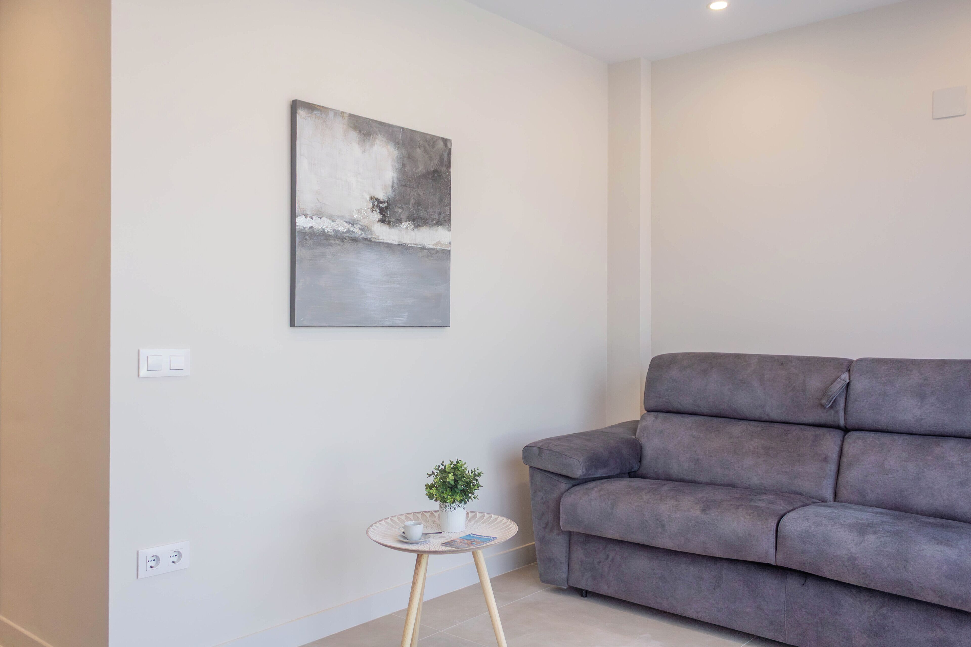 Uma sala de estar moderna com um sofá cinzento, uma mesa redonda pequena com uma planta e um quadro abstrato pendurado na parede.