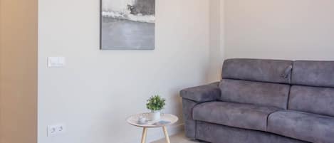 Uma sala de estar moderna com um sofá cinzento, uma mesa redonda pequena com uma planta e um quadro abstrato pendurado na parede.
