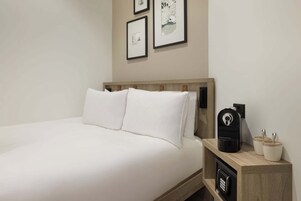 Cama bem arrumada com lençóis brancos, cabeceira de madeira e mesa de cabeceira com cafeteira e uma xícara.