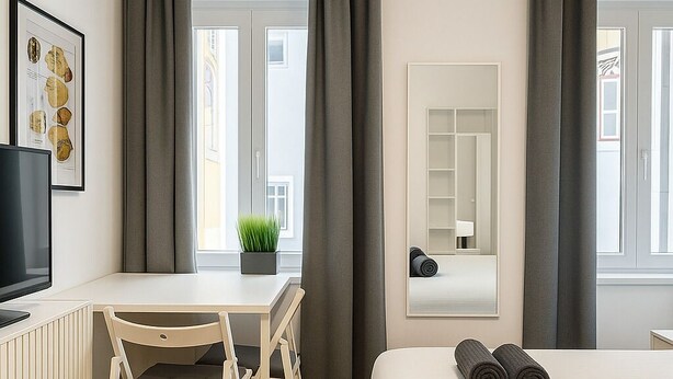 Quarto de hotel com cama, escrivaninha, cadeira, televisão, espelho e janela com cortinas.