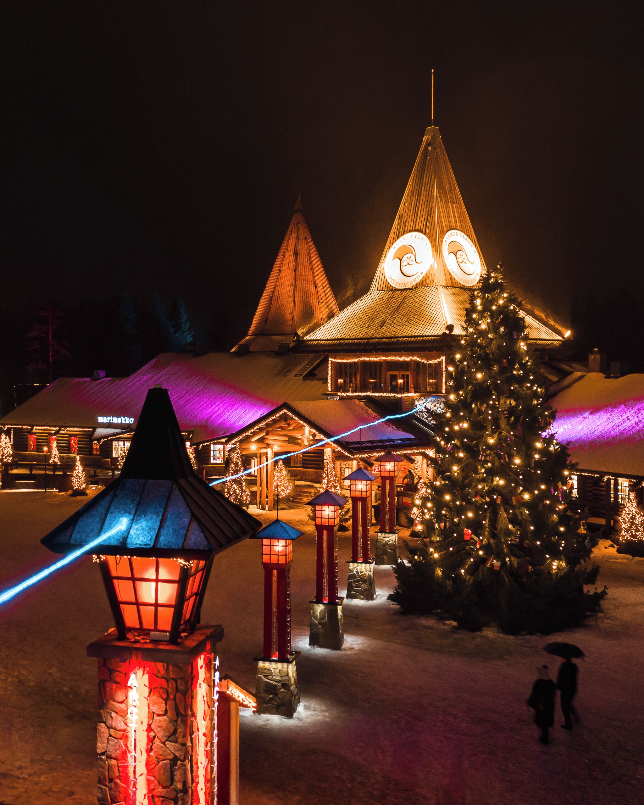 Paisagem coberta de neve com um edifício que se destaca por uma torre e uma árvore de Natal iluminada.