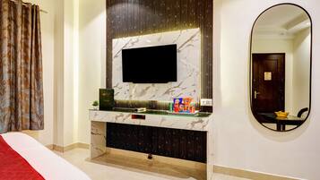 Quarto de hotel com cama, televisão fixada numa parede de mármore, mesa de apoio com petiscos, espelho e porta.