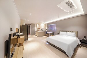 Quarto de hotel moderno com uma cama grande, uma escrivaninha com espelho, um pequeno espaço de cozinha e um banheiro.