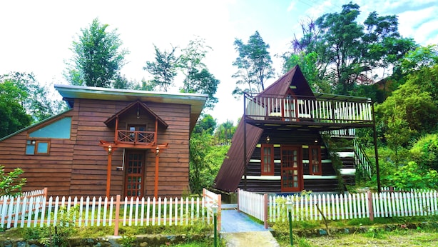 Duas cabanas de madeira com varandas, cercadas por um portão branco e vegetação exuberante.