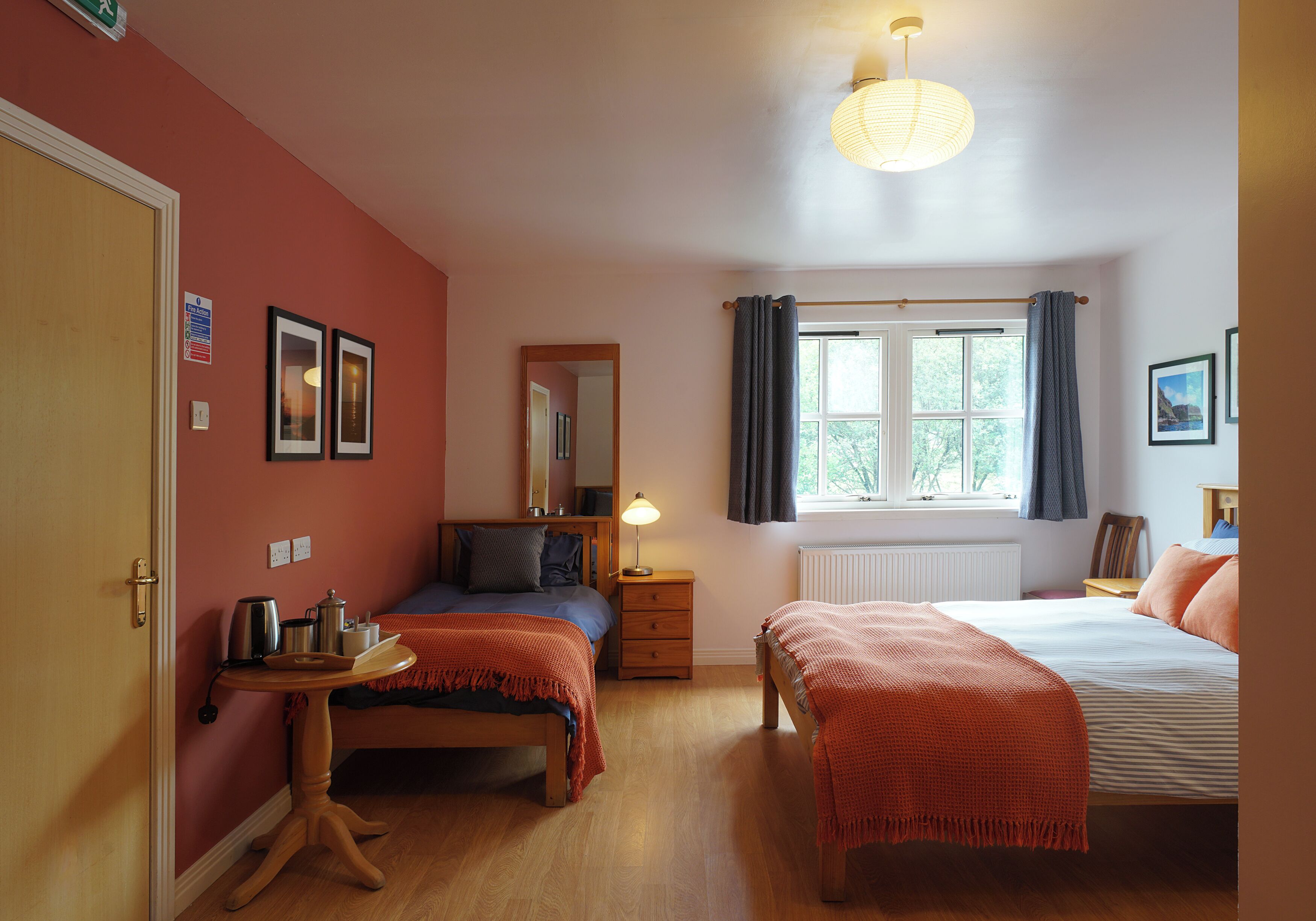 Quarto com duas camas, uma mesa, uma luminária, uma janela com cortinas e um radiador.