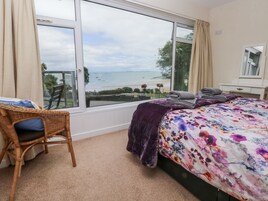 Quarto com uma janela ampla, cama com colcha floral, cadeira de vime e mesa de cabeceira com toalhas.