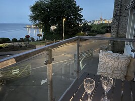 Uma varanda com guarda-corpo de vidro, com vista para uma rua e um corpo de água. Há dois copos de vinho vazios na mesa.