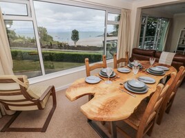 Uma sala de jantar com vista para o mar, mesa de madeira posta com pratos e copos, e cadeiras de couro.