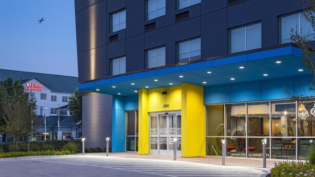 Entrada moderna de um hotel com fachada azul e amarela, com a placa Hilton Garden Inn visível, e um estacionamento em frente.